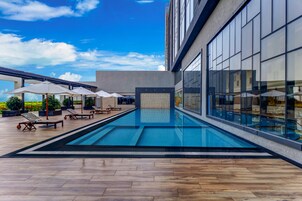 Uma área de piscina no terraço com espreguiçadeiras e guarda-sóis, cercada por paredes de vidro e um céu limpo.
