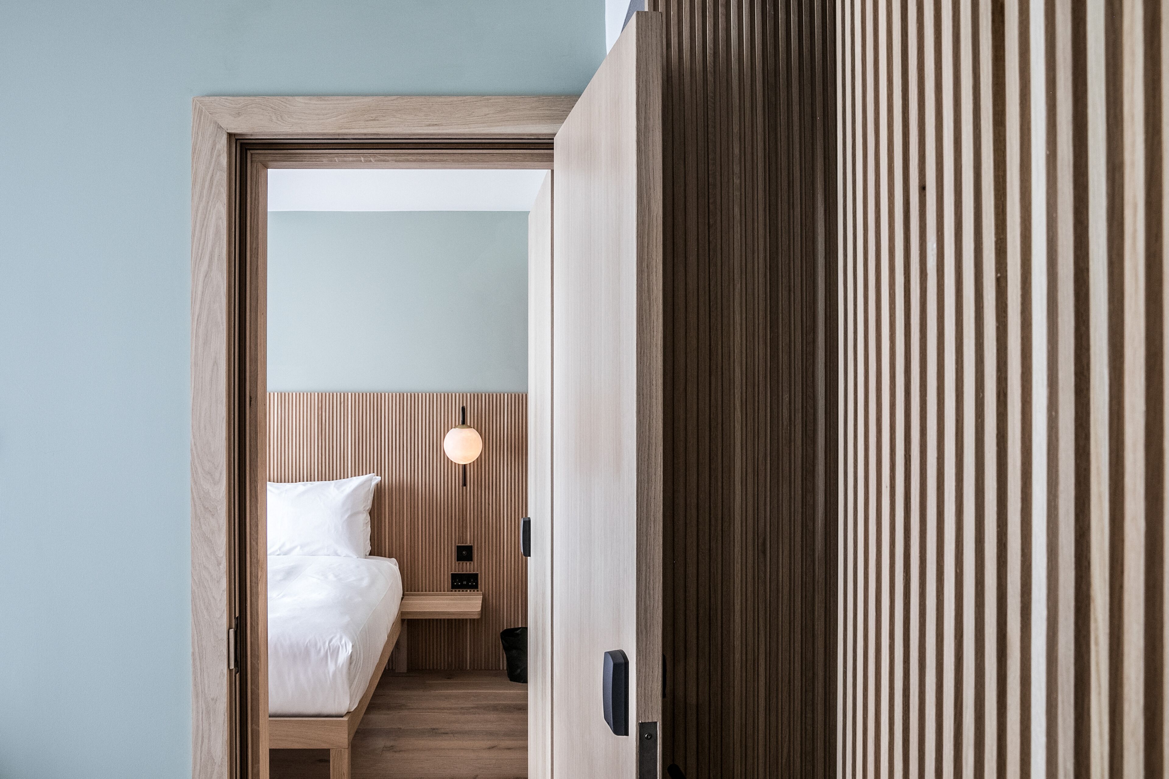 Quarto de hotel moderno com uma cama de madeira, uma mesa de cabeceira, um abajur e um guarda-roupa de madeira.