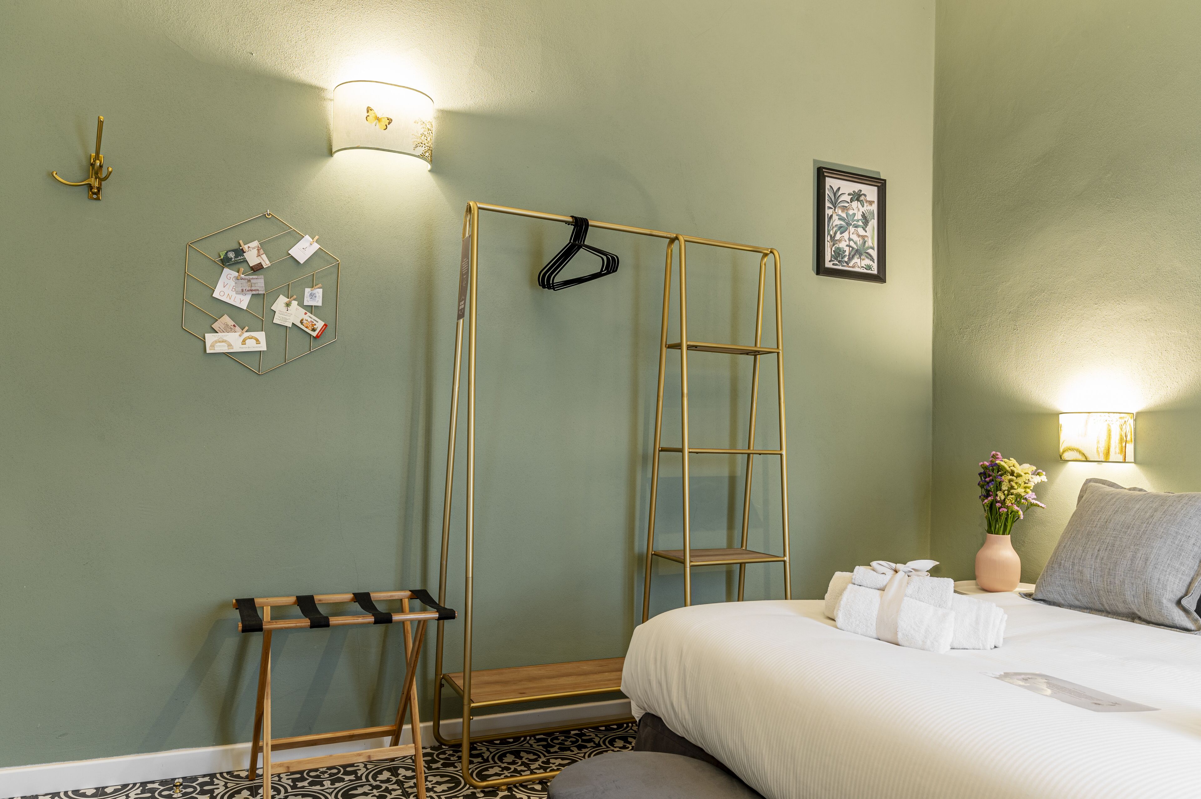 Quarto de hotel bem arrumado com uma cama, um cabideiro, um pequeno banco e um candeeiro fixado na parede.