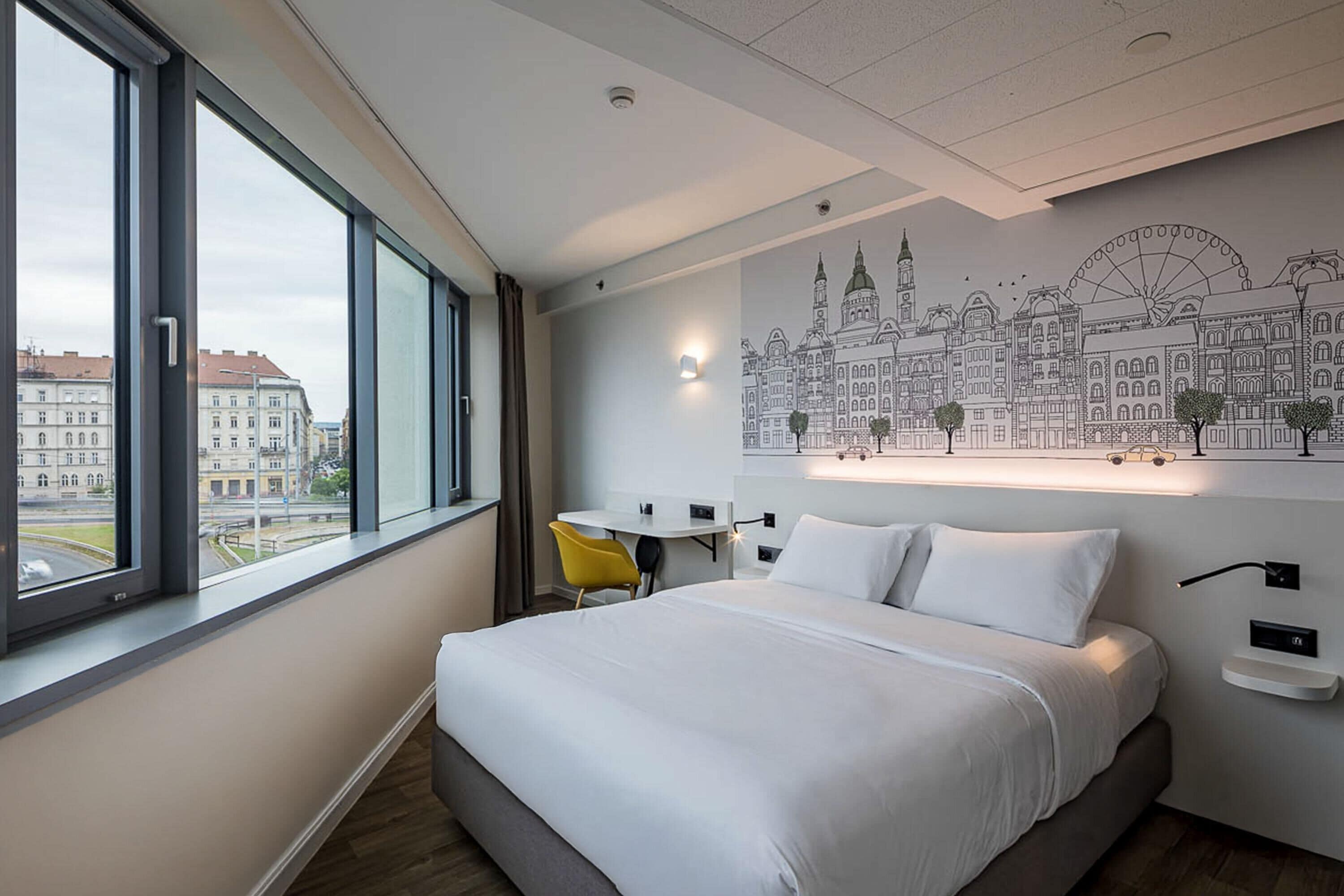 Quarto de hotel com uma cama grande, uma escrivaninha, uma cadeira e um mural com vista para a cidade na parede.