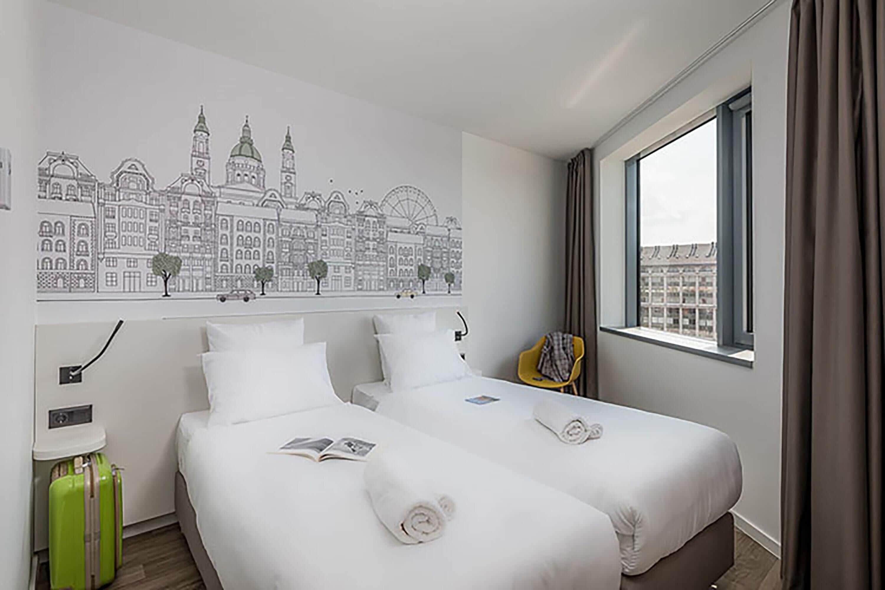 Quarto de hotel com duas camas, um mural da paisagem urbana, uma janela com cortinas e uma mala.