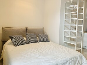 Uma cama bem arrumada com roupa de cama branca e três almofadas estampadas, cabeceira bege e guarda-roupa branco com espaço para pendurar.