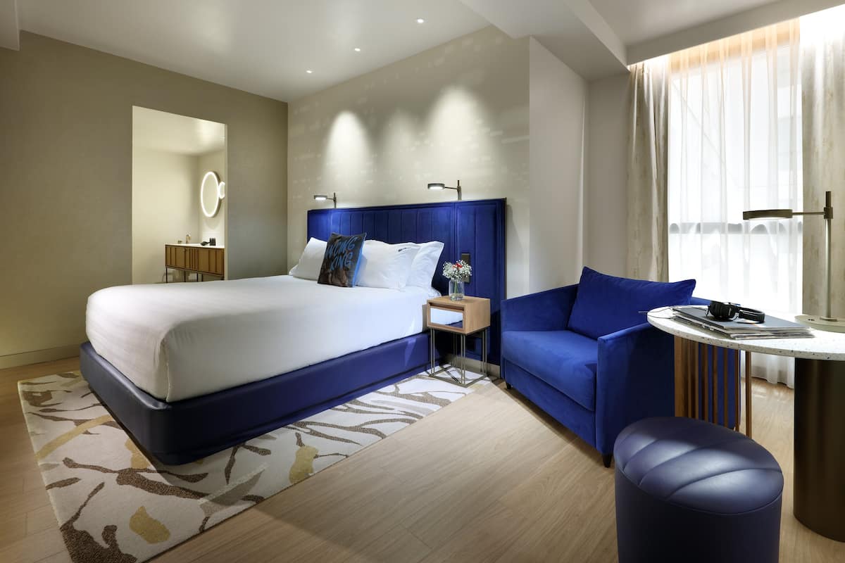 Quarto de hotel moderno com uma cama grande, um sofá azul, uma mesinha e um espelho.