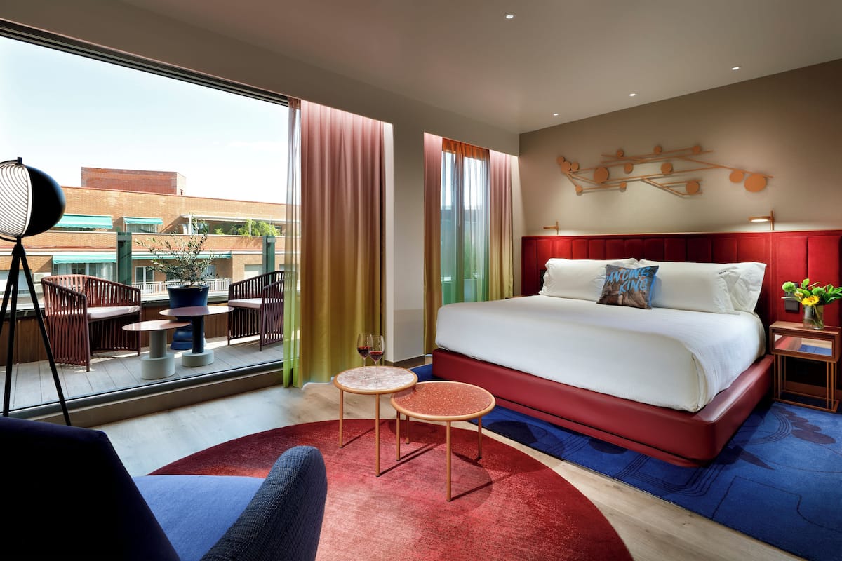 Quarto de hotel moderno com uma cama grande, uma mesa pequena, uma cadeira e vista da cidade por grandes janelas.