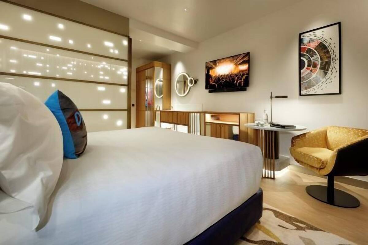 Quarto de hotel com uma cama grande, uma mesa com cadeira, televisão fixada na parede e uma obra de arte abstrata emoldurada.