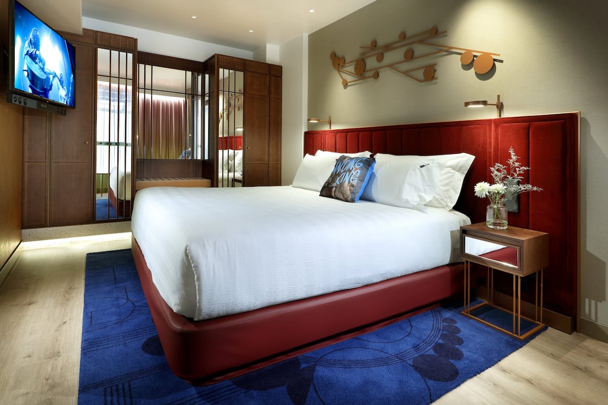Quarto de hotel com uma cama grande, uma TV fixada na parede e cabeceira vermelha.