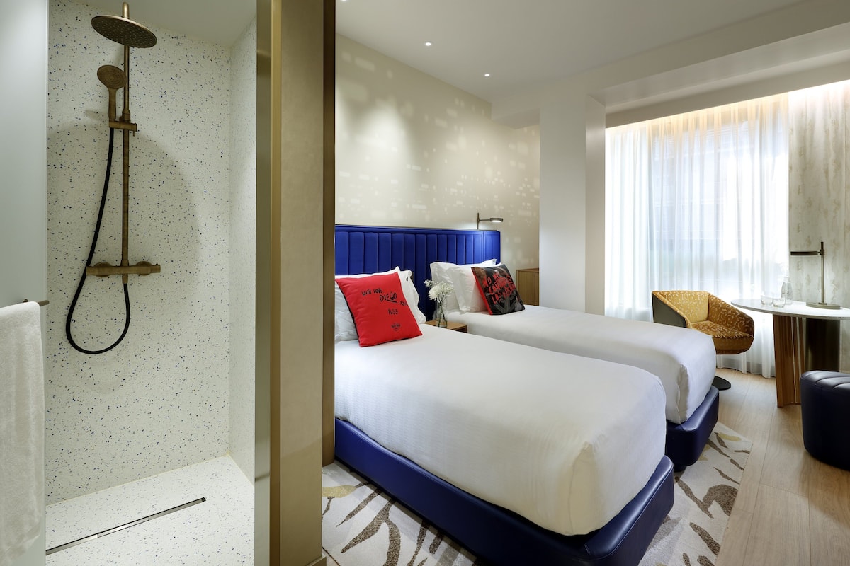 Quarto de hotel com duas camas, chuveiro, uma mesinha e uma cadeira.