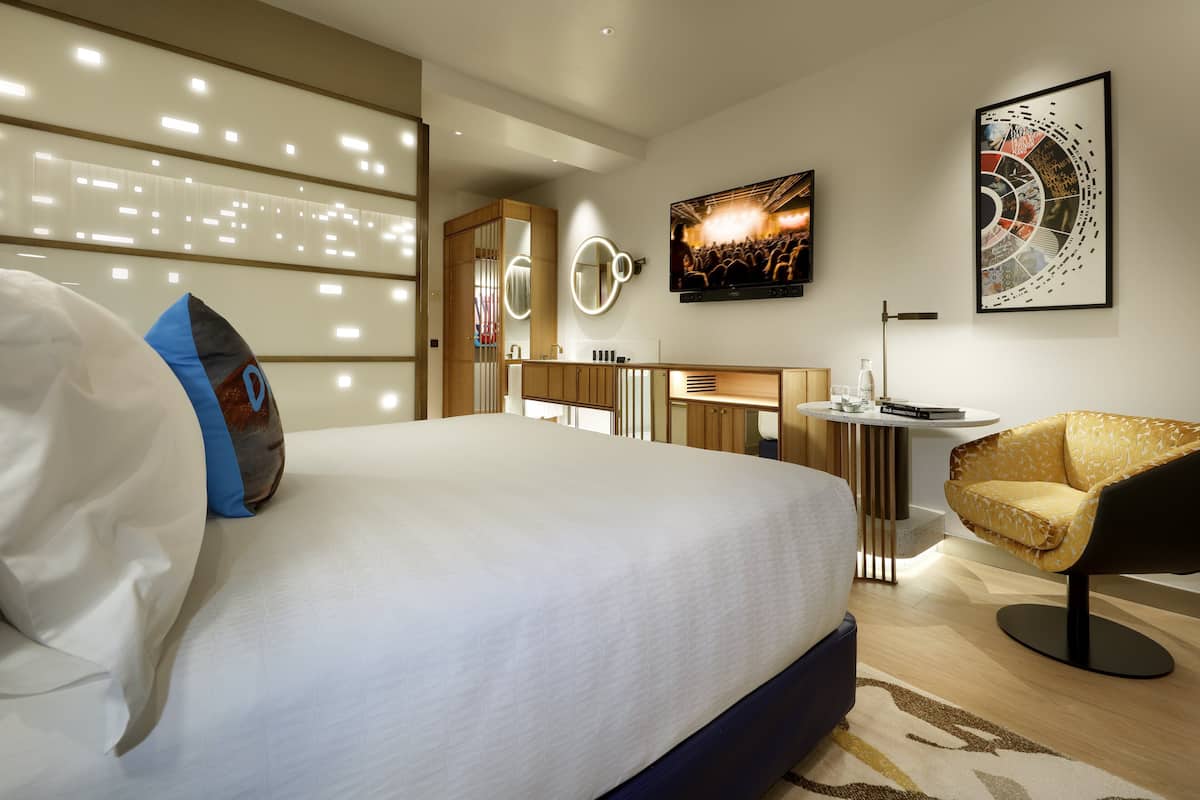 Quarto de hotel moderno com uma cama grande, uma escrivaninha, uma cadeira e uma televisão na parede.