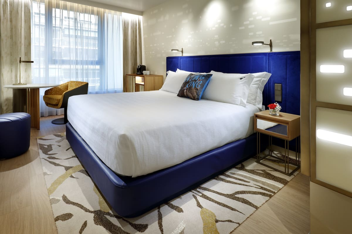 Quarto de hotel moderno com uma cama grande, uma escrivaninha, uma cadeira e uma mesa de cabeceira com abajur.