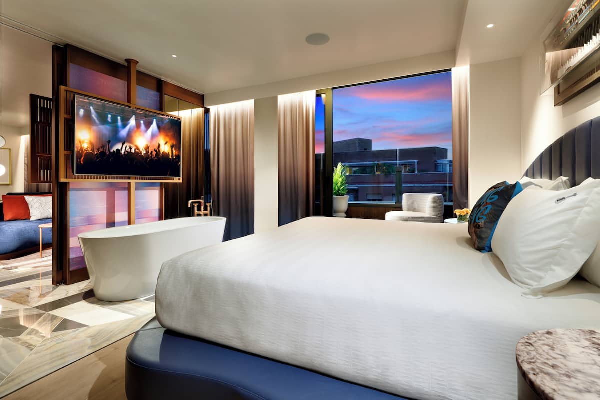 Quarto de hotel moderno com uma cama grande, uma banheira, uma televisão de tela plana e vista para a paisagem urbana.