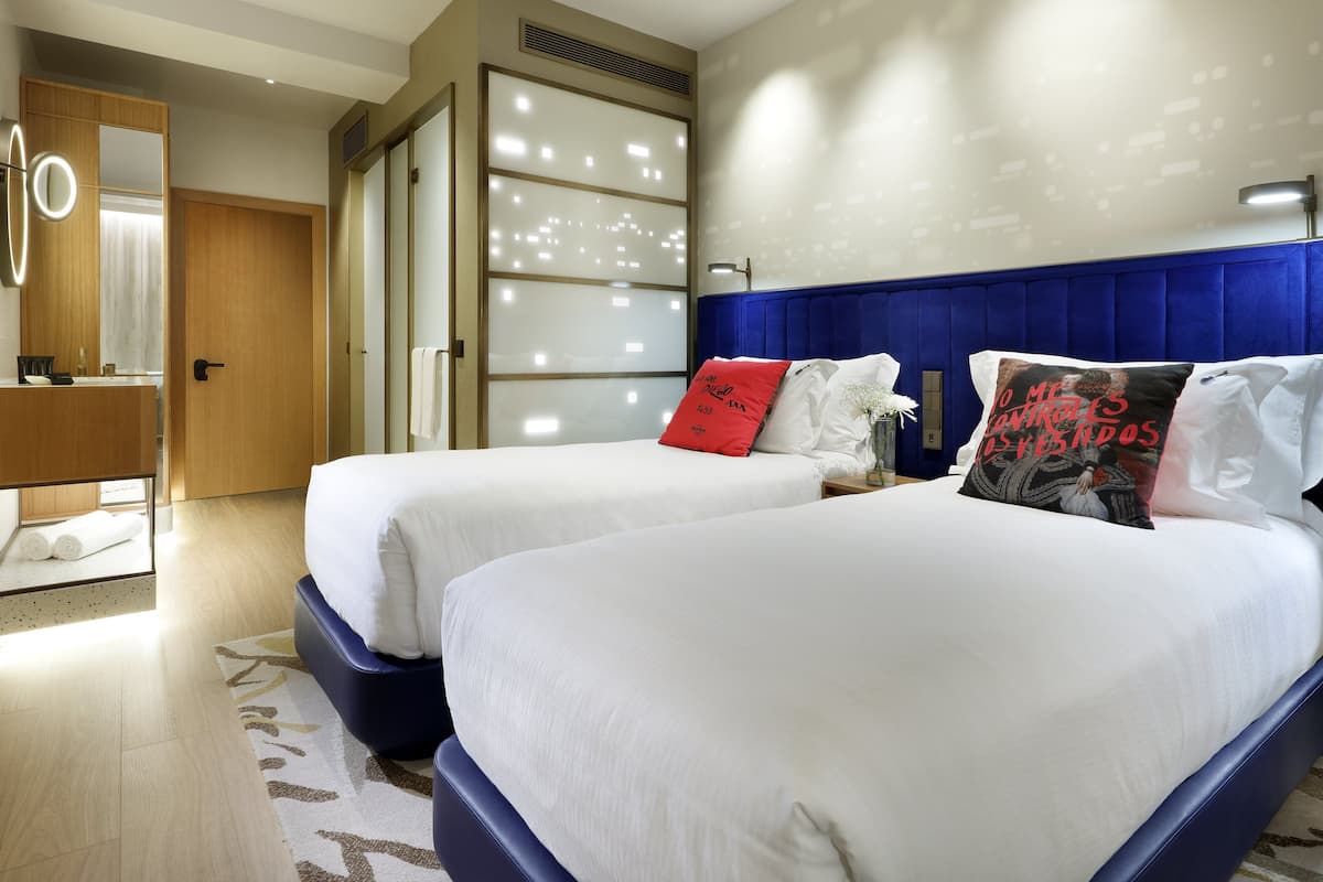 Quarto de hotel com duas camas, um guarda-roupa de madeira, um espelho e banheiro com porta de vidro.