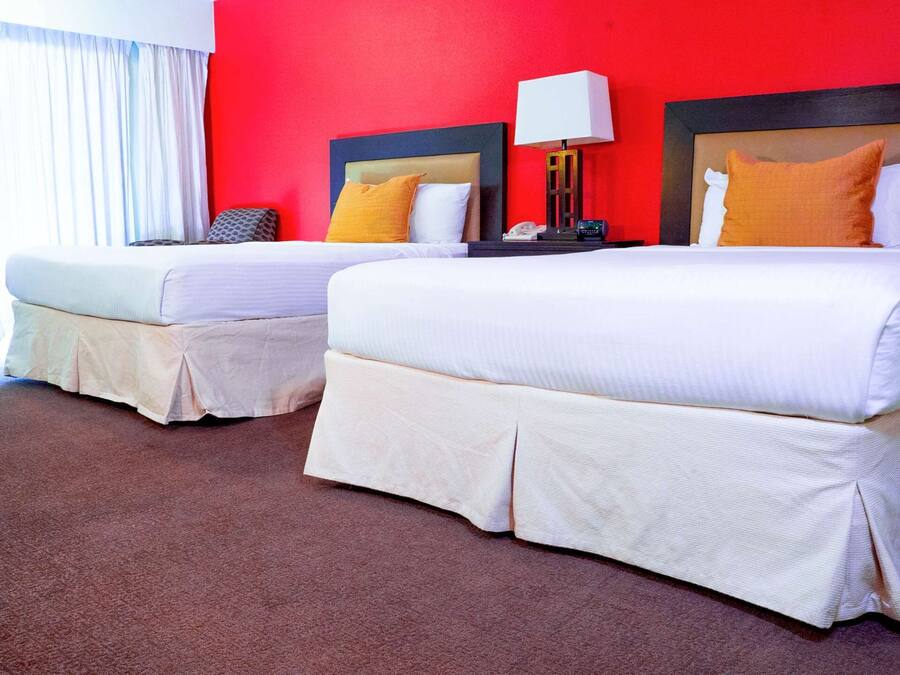 Duas camas com roupa de cama branca, uma parede vermelha e uma luminária branca sobre uma mesa de cabeceira.