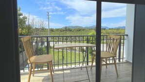 Uma varanda com mesa e cadeiras de madeira, grade de metal e vista para uma paisagem verde com árvores e colinas.