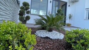 Uma área de recepção com um edifício branco, entrada pavimentada e um jardim ajardinado com pedras e arbustos.