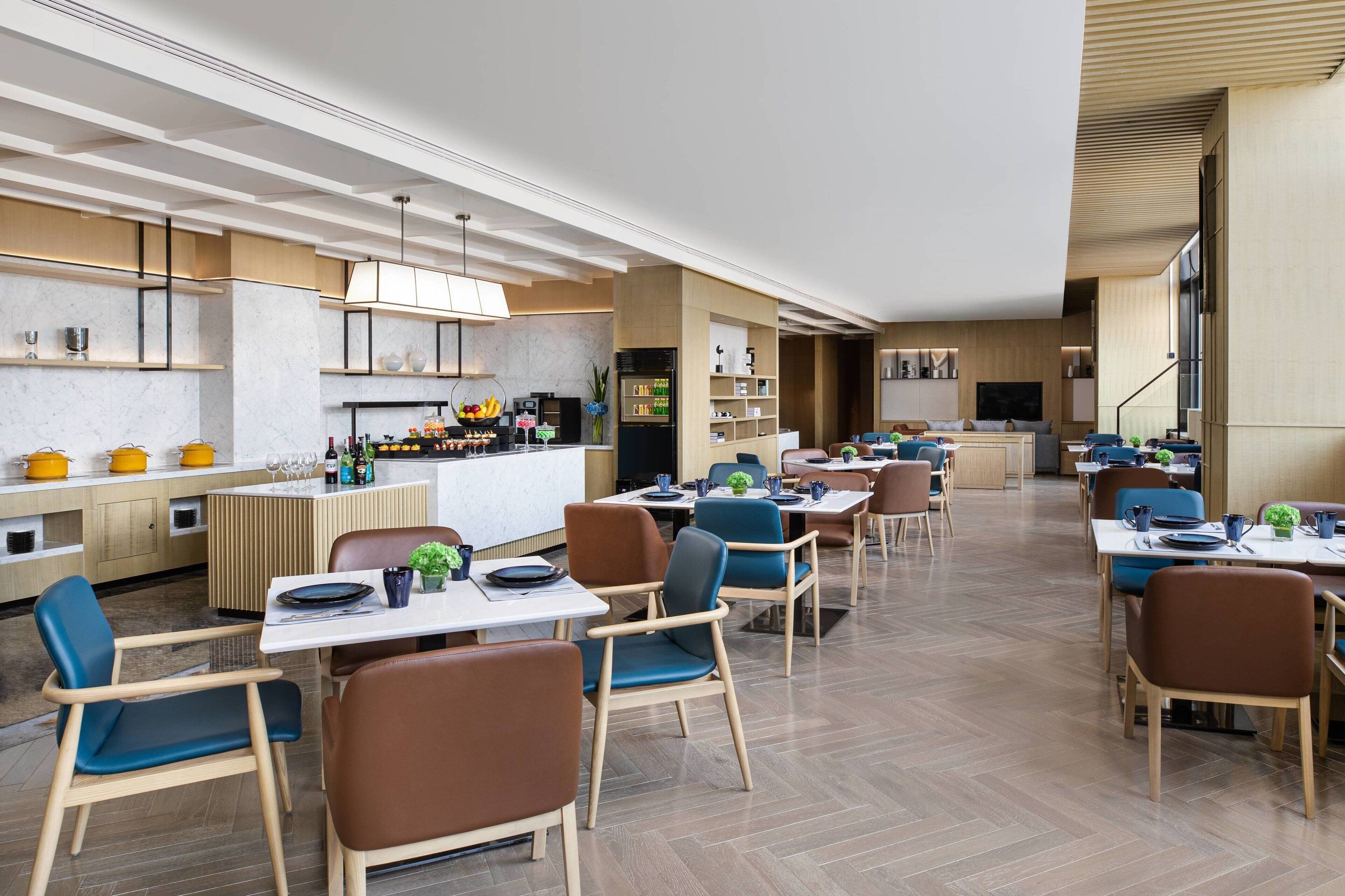 Uma área de refeições moderna com várias mesas e cadeiras, uma cozinha com balcão e uma sala de estar com televisão.