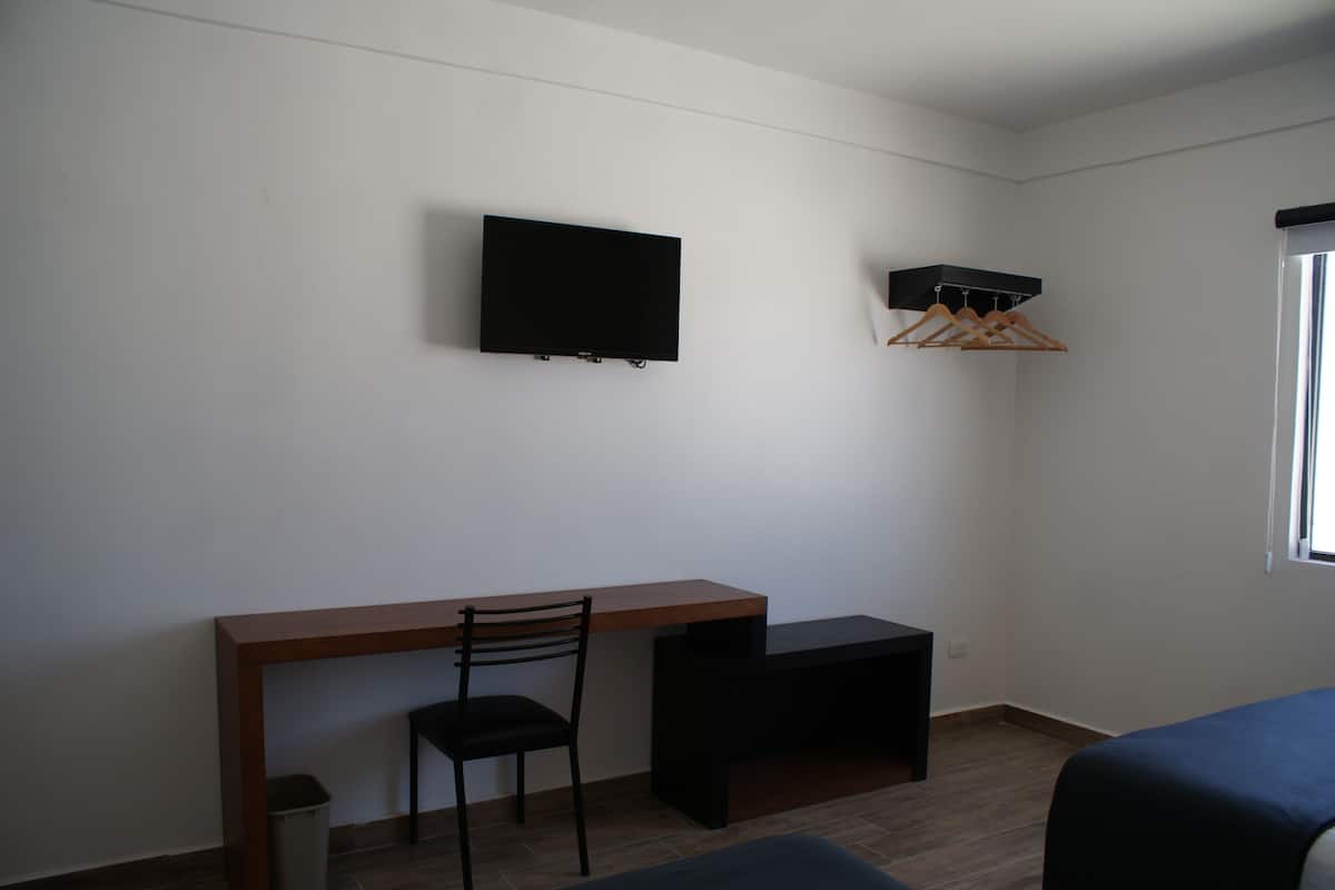 Quarto pequeno e pouco mobiliado, com uma escrivaninha, cadeira e televisão fixada na parede.