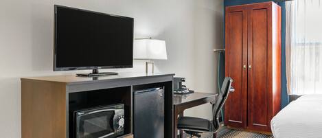 Quarto de hotel com televisão de tela plana, mini-geladeira, micro-ondas, escrivaninha com luminária, cadeira, armário e cama.