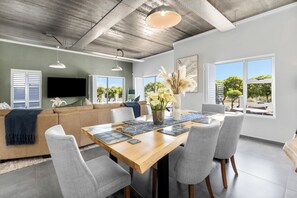 Sala de estar moderna com uma mesa de jantar de madeira, cadeiras estofadas em cinza, um sofá bege e uma grande janela com vista para vegetação.