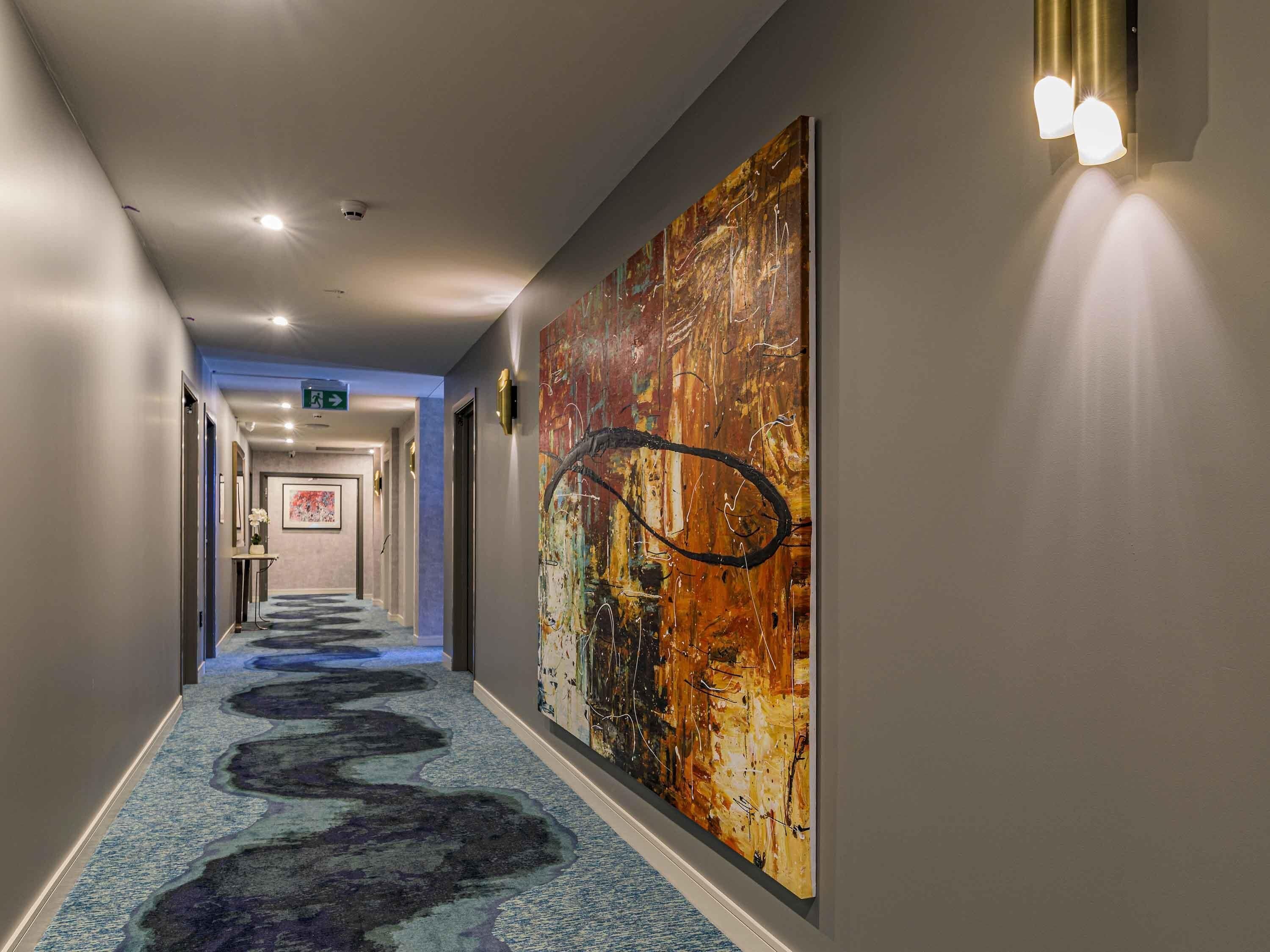 Corredor de hotel com uma grande pintura abstrata na parede direita, um tapete estampado e um aparelho de iluminação acima da pintura.