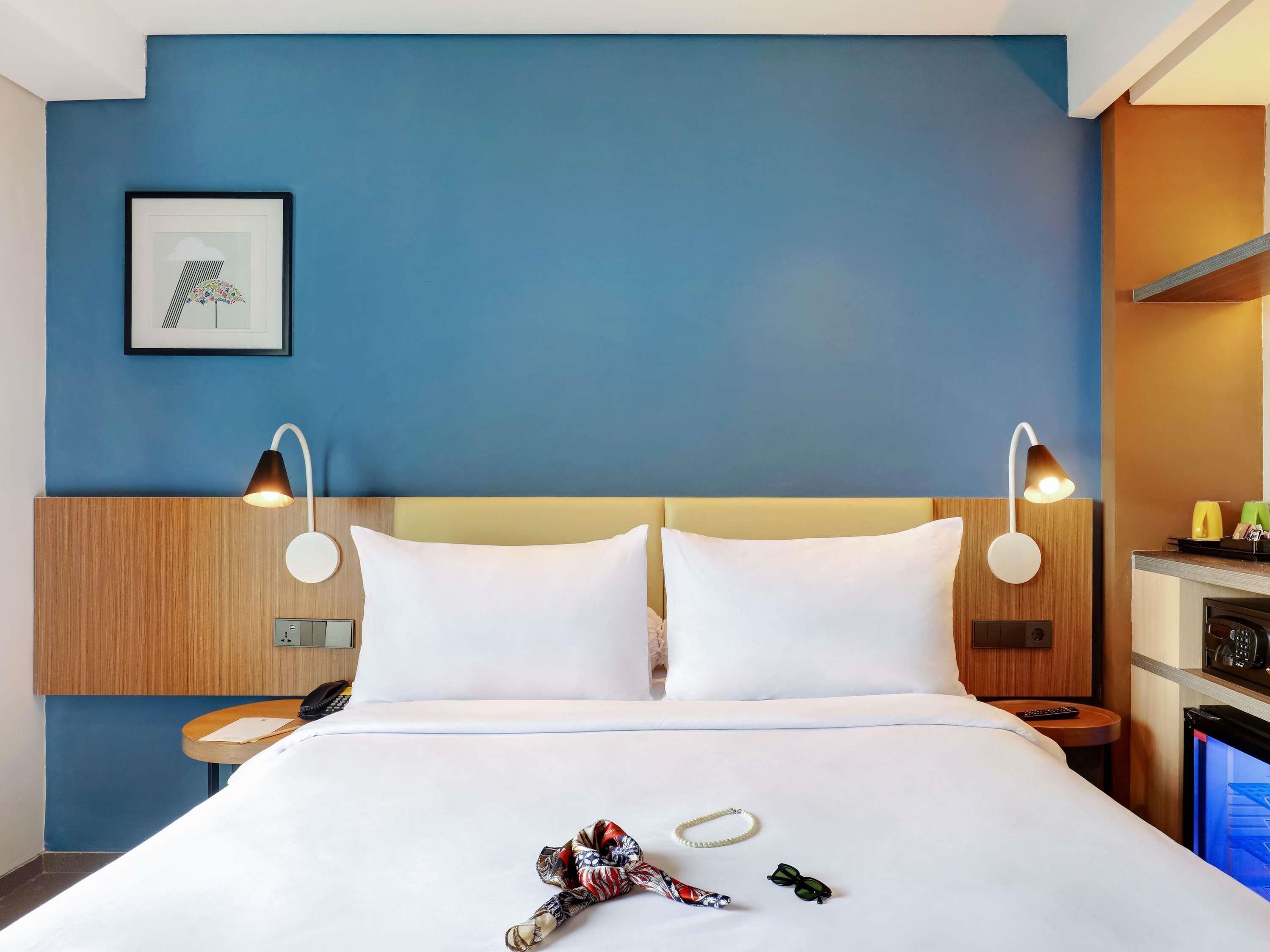Quarto de hotel com parede azul, cabeceira de madeira, duas luminárias de apoio, cama com roupa branca, quadro emoldurado na parede, mesinha com telefone e televisão no lado direito.
