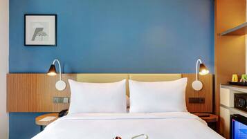 Quarto de hotel com parede azul, cabeceira de madeira, duas luminárias de apoio, cama com roupa branca, quadro emoldurado na parede, mesinha com telefone e televisão no lado direito.