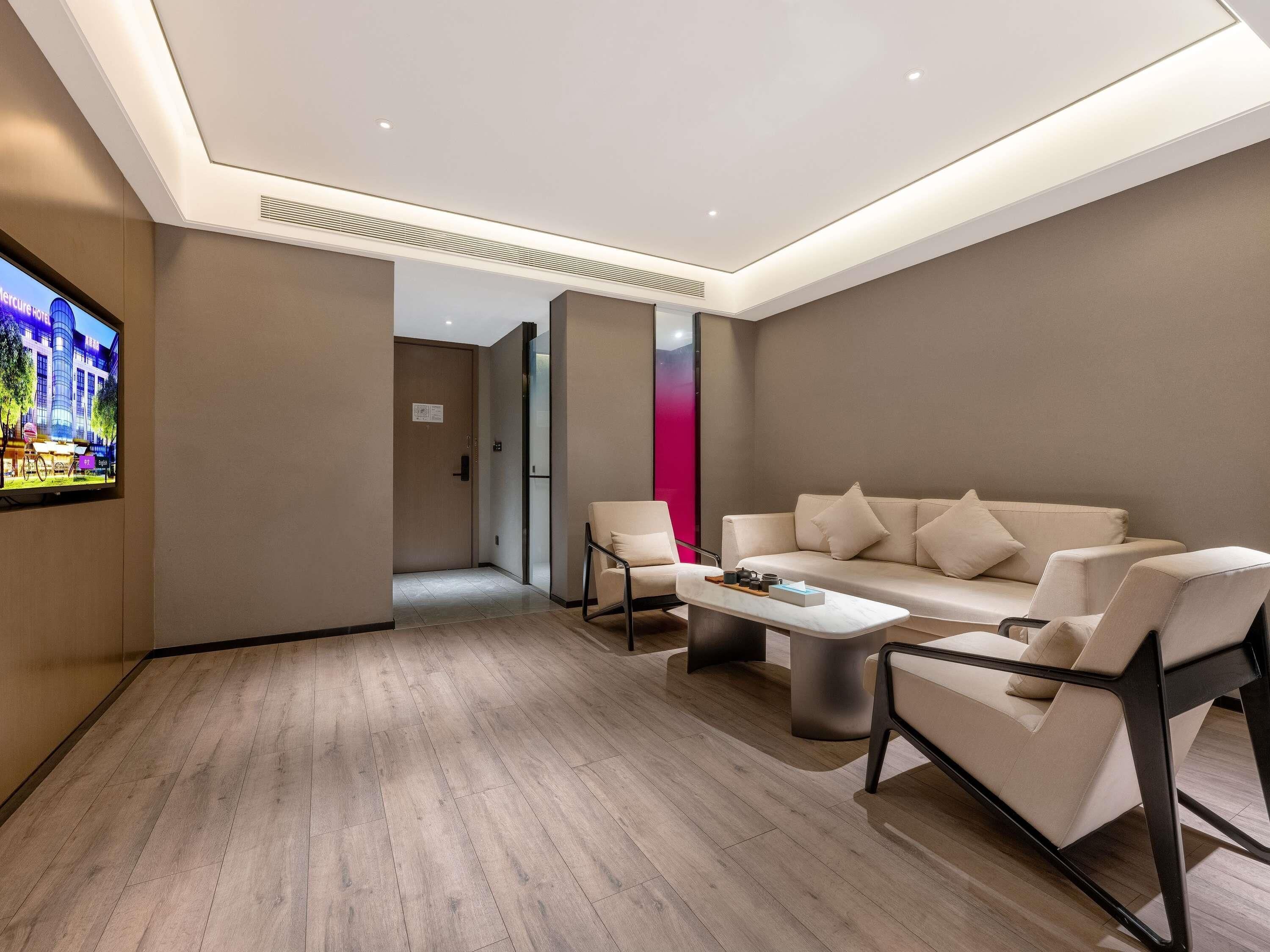 Quarto de hotel moderno com sofá, poltronas, mesa de centro e TV de tela plana fixada na parede.