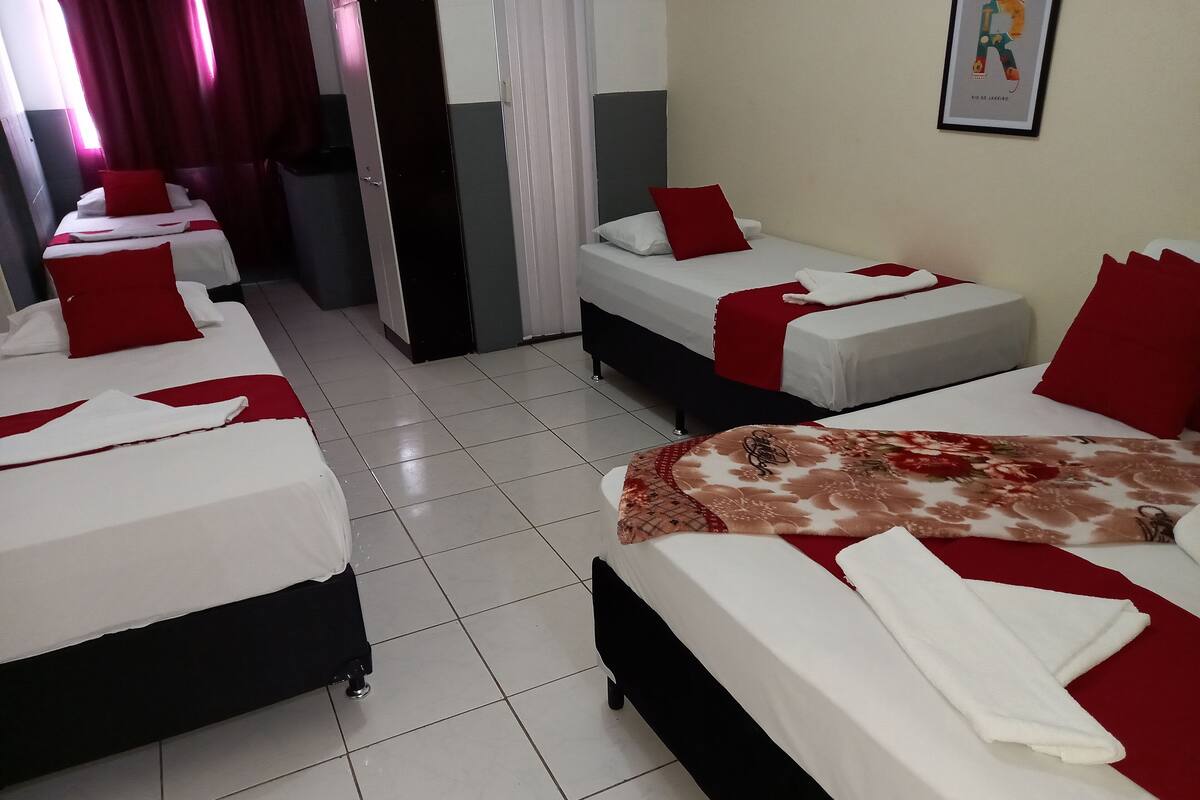Quarto de hotel com três camas, cada uma com roupa de cama branca e almofadas vermelhas. O quarto possui um chão de azulejos e um quadro emoldurado na parede.