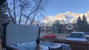 Uma banheira de hidromassagem com uma pessoa sentada, com vista para uma cordilheira nevada e um carro vermelho estacionado na rua.