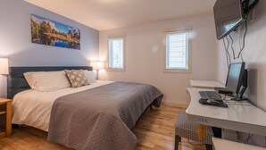 Um quarto com cama, luminárias de cabeceira, escrivaninha com computador e uma janela.