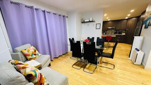 Sala de estar moderna com um sofá cinzento, almofadas coloridas, uma mesa de jantar com cadeiras pretas e uma área de cozinha ao fundo.
