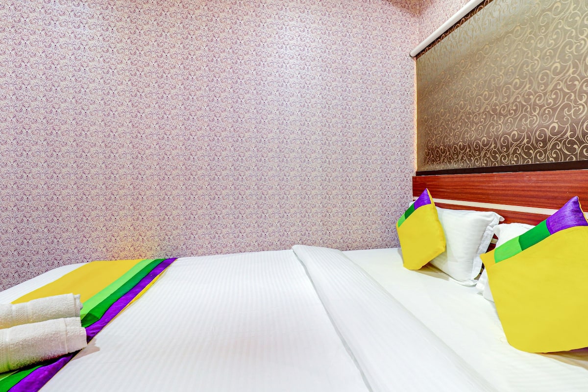 Quarto de hotel com papel de parede estampado, cabeceira de madeira e cama bem arrumada com almofadas coloridas e manta listrada.