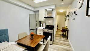 Cozinha compacta com geladeira, fogão e micro-ondas. Há uma mesa de jantar com cadeiras e uma pequena planta sobre ela. A cozinha possui piso claro e paredes brancas.
