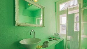 Um banheiro com parede verde, pia branca, espelho e janela com vista para o exterior.