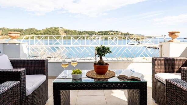 Terraço com móveis de vime, mesa de vidro, planta em vaso e vista para o mar.