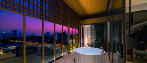 Interior moderno com uma grande janela de vidro, banheira de hidromassagem e vista para a cidade ao entardecer.