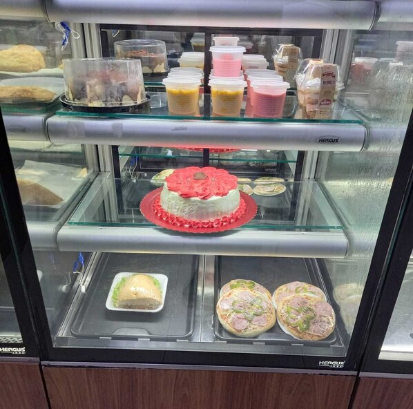 Vitrine de uma padaria com diversos produtos de panificação, incluindo um bolo com cobertura vermelha, sanduíches e doces.