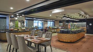 Restaurante com serviço de buffet, incluindo um balcão com diversos pratos e uma área de refeições com mesas e cadeiras.