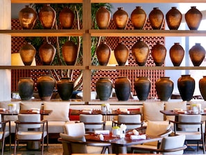 Um restaurante com várias mesas e cadeiras, uma variedade de vasos em prateleiras, e uma parede estampada ao fundo.