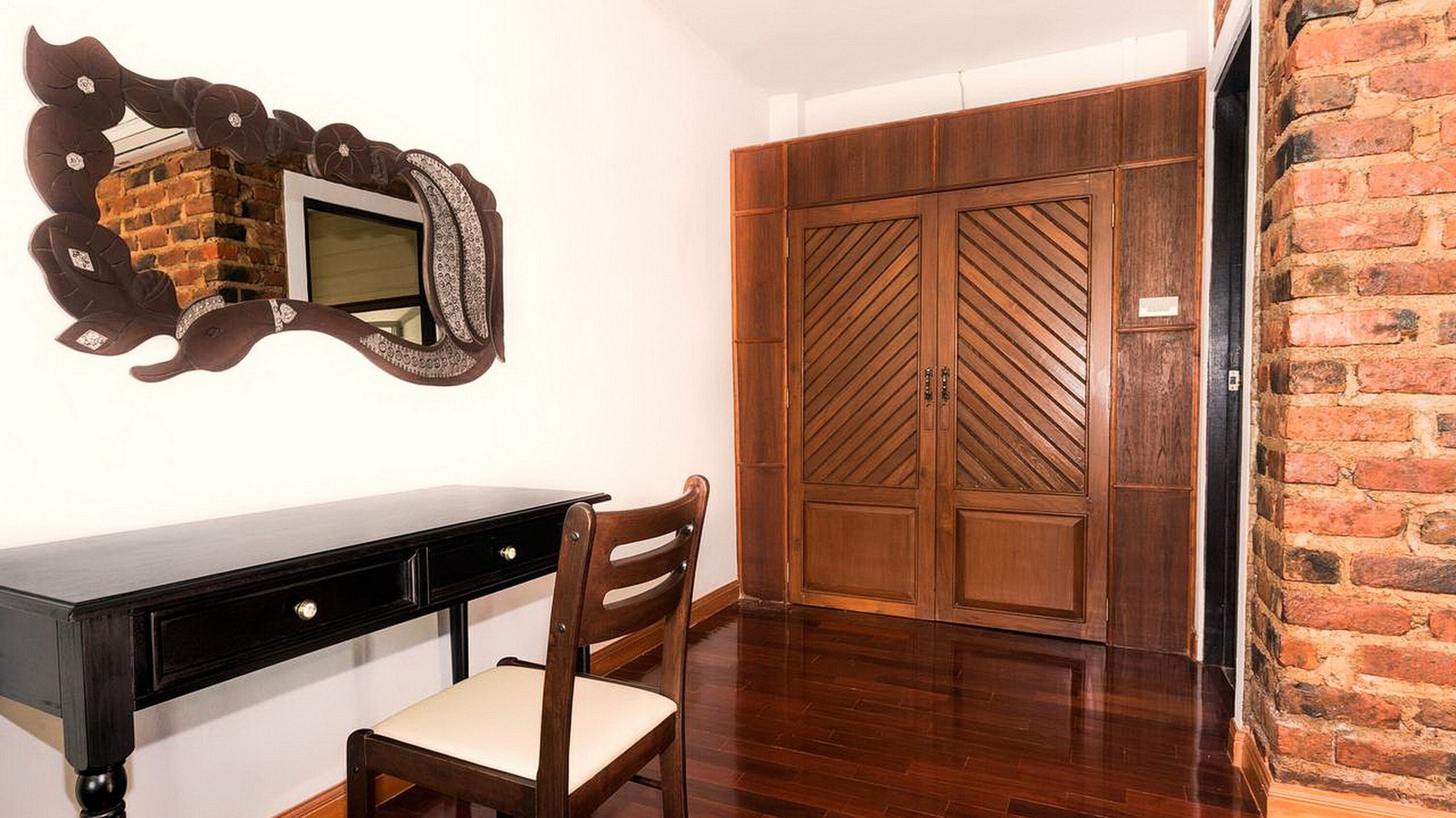 Quarto com escrivaninha de madeira, uma cadeira, um espelho com moldura decorativa e um guarda-roupa de madeira duplo.