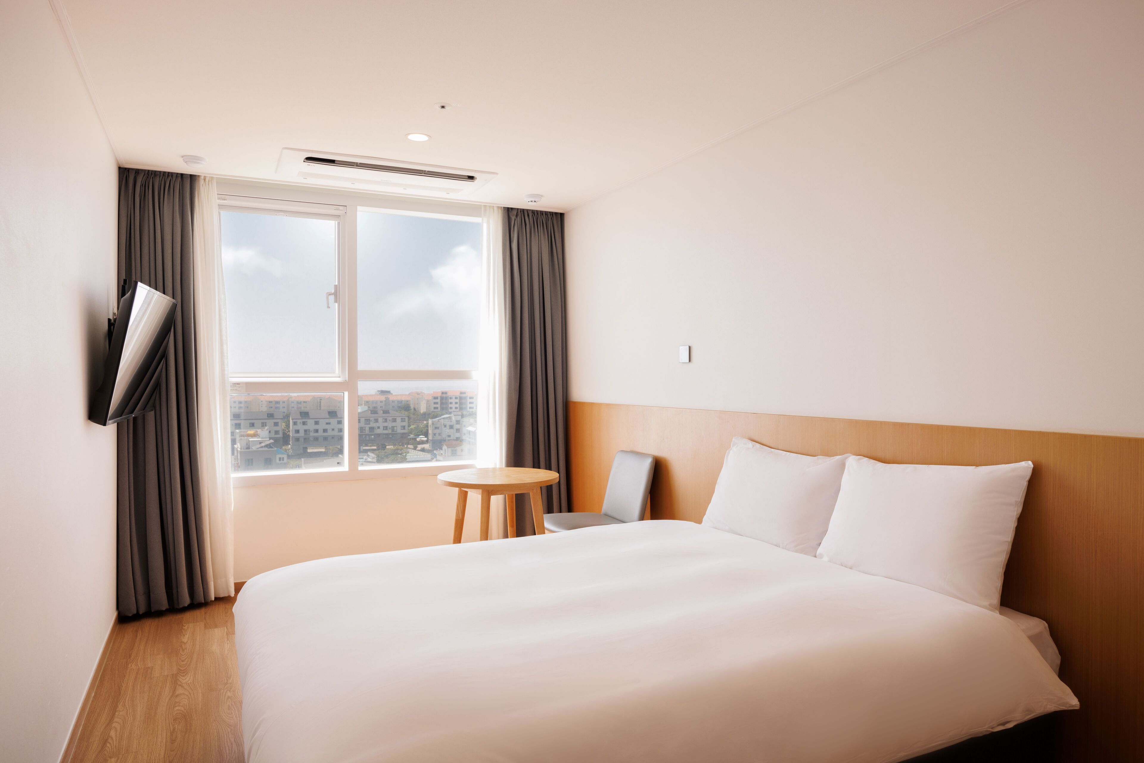 Quarto de hotel com uma cama grande, uma mesa redonda pequena, uma cadeira, uma televisão fixada na parede e uma janela com cortinas.