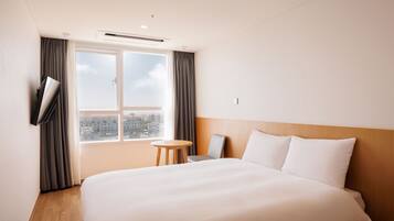 Quarto de hotel com uma cama grande, uma mesa redonda pequena, uma cadeira, uma televisão fixada na parede e uma janela com cortinas.