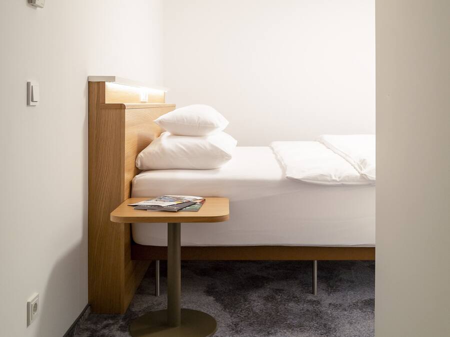 Um quarto pequeno e bem arrumado com uma cama de solteiro, uma mesa de cabeceira com uma revista e uma cabeceira de madeira.
