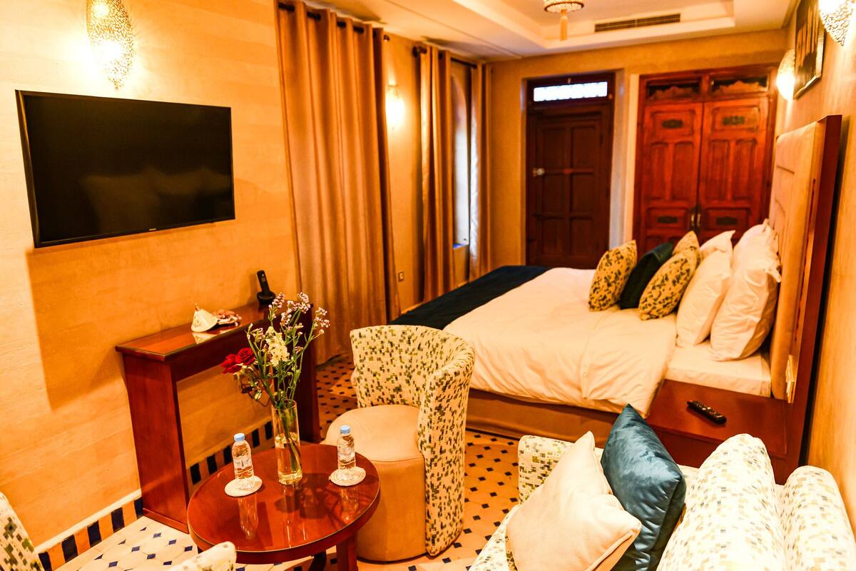 Quarto de hotel com cama, televisão na parede, uma mesinha com um vaso de flores e uma cadeira.