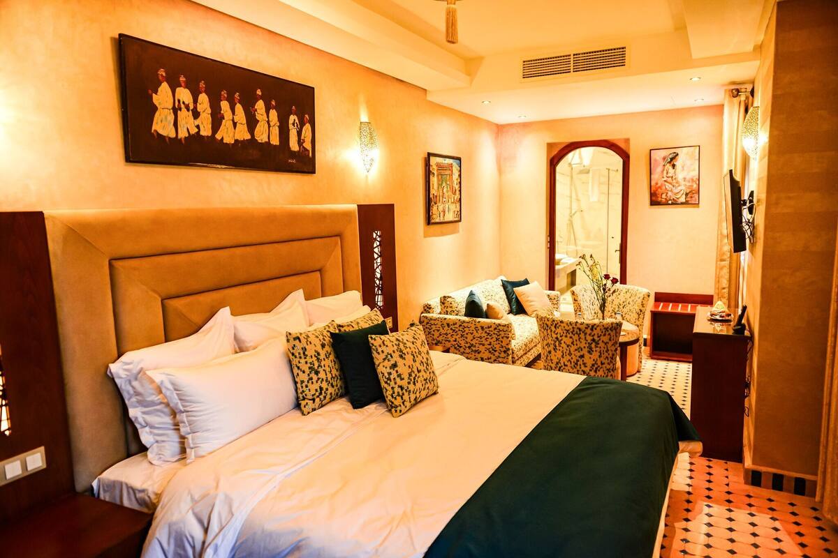Quarto de hotel com uma cama grande, um espaço de estar com sofá e poltronas, um espelho e obras de arte nas paredes.