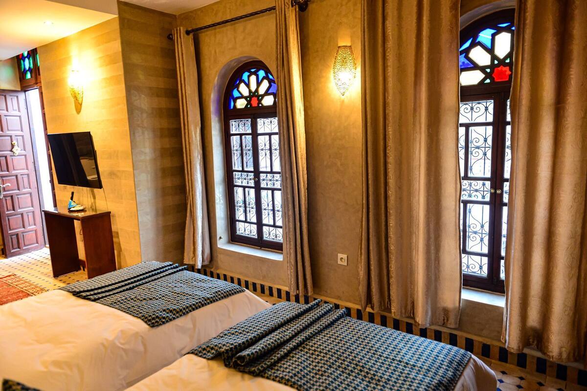 Quarto de hotel com duas camas, um espelho, uma televisão e duas janelas com grades de ferro decorativas.