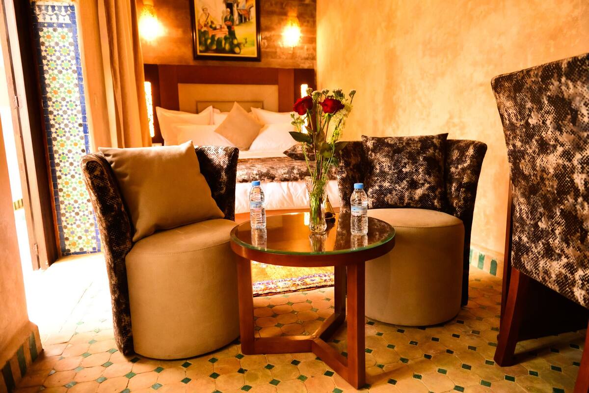Quarto de hotel com uma cama, duas poltronas, uma mesinha com garrafas de água e um vaso com flores.