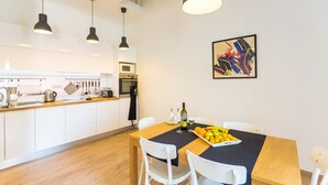 Cozinha moderna com armários brancos, bancada de madeira e luminárias pendentes pretas. Uma mesa de jantar com cadeiras brancas e um jogo americano preto, disposta com uma garrafa, copos e frutas.