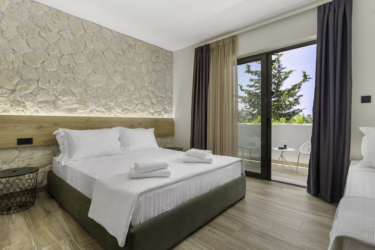 Quarto de hotel moderno com parede de destaque em pedra, uma cama grande com roupa de cama branca, cabeceira de madeira e uma varanda com vista para árvores.