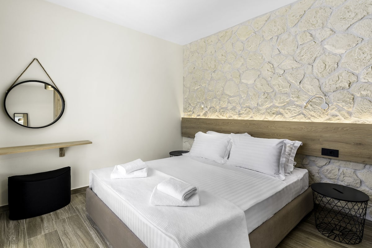 Quarto de hotel moderno com parede de pedra, espelho redondo, cabeceira de madeira, cama bem arrumada com roupa de cama branca, um banquinho preto e uma pequena mesa lateral redonda.