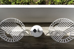 Uma mesa redonda com duas cadeiras brancas, um objeto preto sobre a mesa e uma parede branca com vegetação ao fundo.